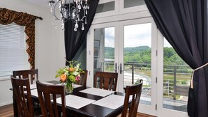 Uma sala de jantar com uma mesa posta para quatro pessoas, um vaso com flores e vista para uma paisagem verde através de grandes portas de vidro.