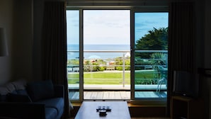Quarto com sofá, mesa de centro e televisão. Possui uma grande porta de vidro deslizante que dá para uma varanda com vista para o mar e um jardim.