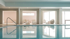 Piscina interior moderna com água cristalina, rodeada por espreguiçadeiras brancas e um ambiente iluminado com amplas janelas.