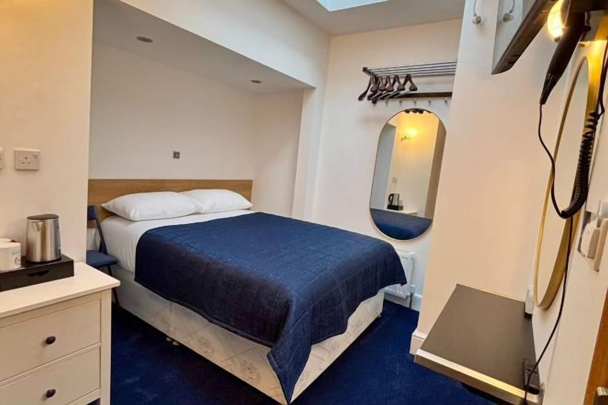 Quarto de hotel com cama, mesa de cabeceira com chaleira, espelho e pequena mesa com um laptop.
