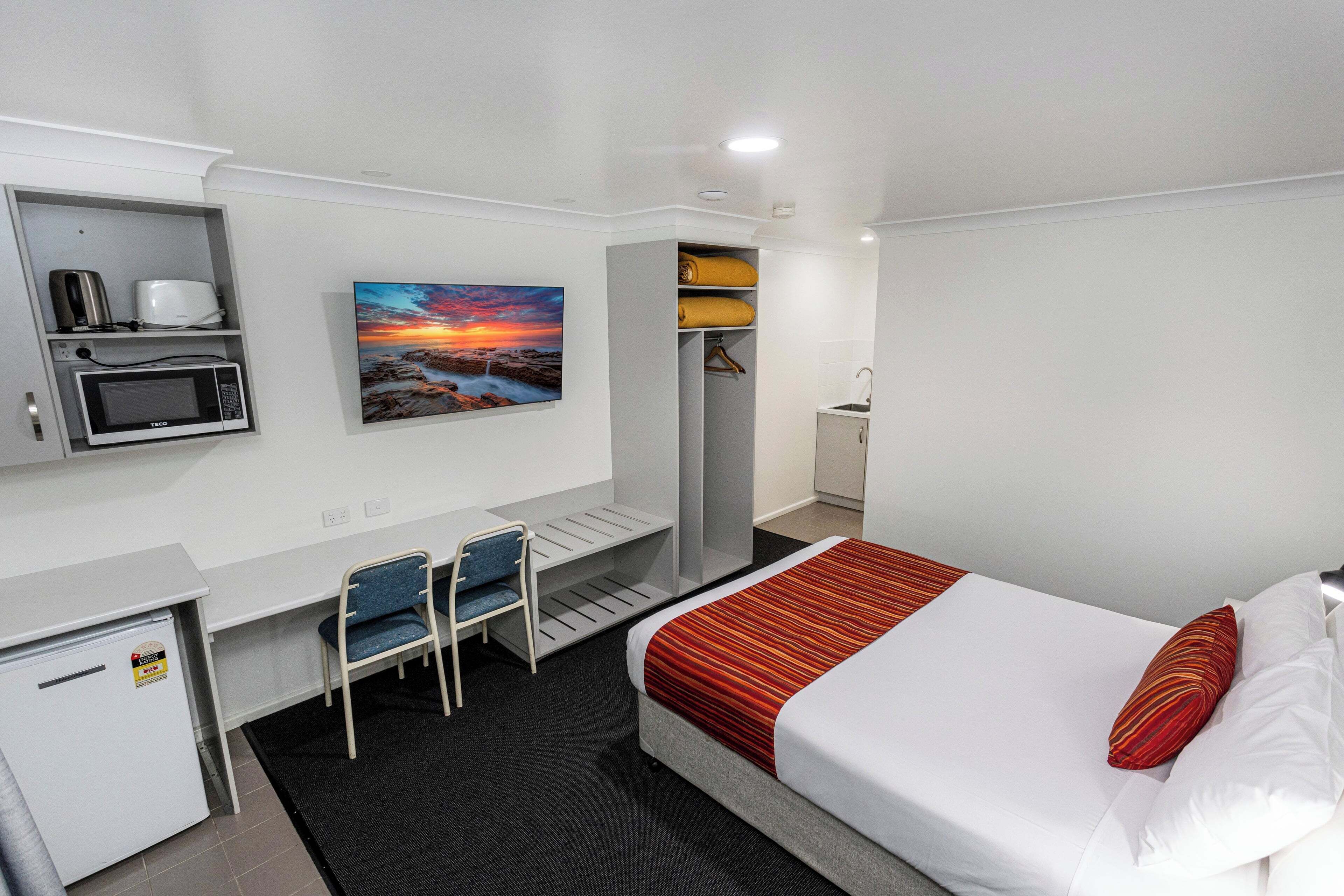 Quarto de hotel com cama, escrivaninha, cadeira, micro-ondas, mini-geladeira, quadro e guarda-roupa.
