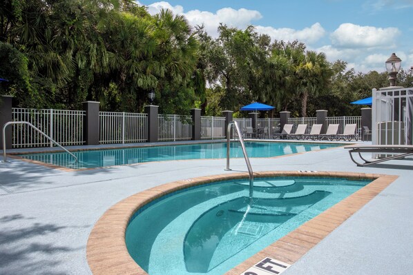 Piscina com uma pequena banheira de hidromassagem, rodeada por espreguiçadeiras e guarda-sóis, além de uma cerca e árvores ao fundo.