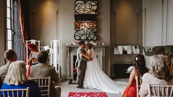Cerimônia de casamento com um casal se beijando em frente a uma lareira, convidados sentados em cadeiras, e um tapete vermelho com pétalas de flores.
