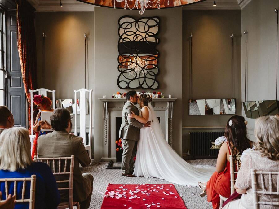 Cerimônia de casamento com um casal se beijando em frente a uma lareira, convidados sentados em cadeiras, e um tapete vermelho com pétalas de flores.