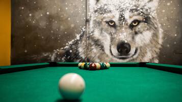 Uma mesa de bilhar com bola branca e bolas listradas, um mural de lobo ao fundo e superfície de feltro verde.