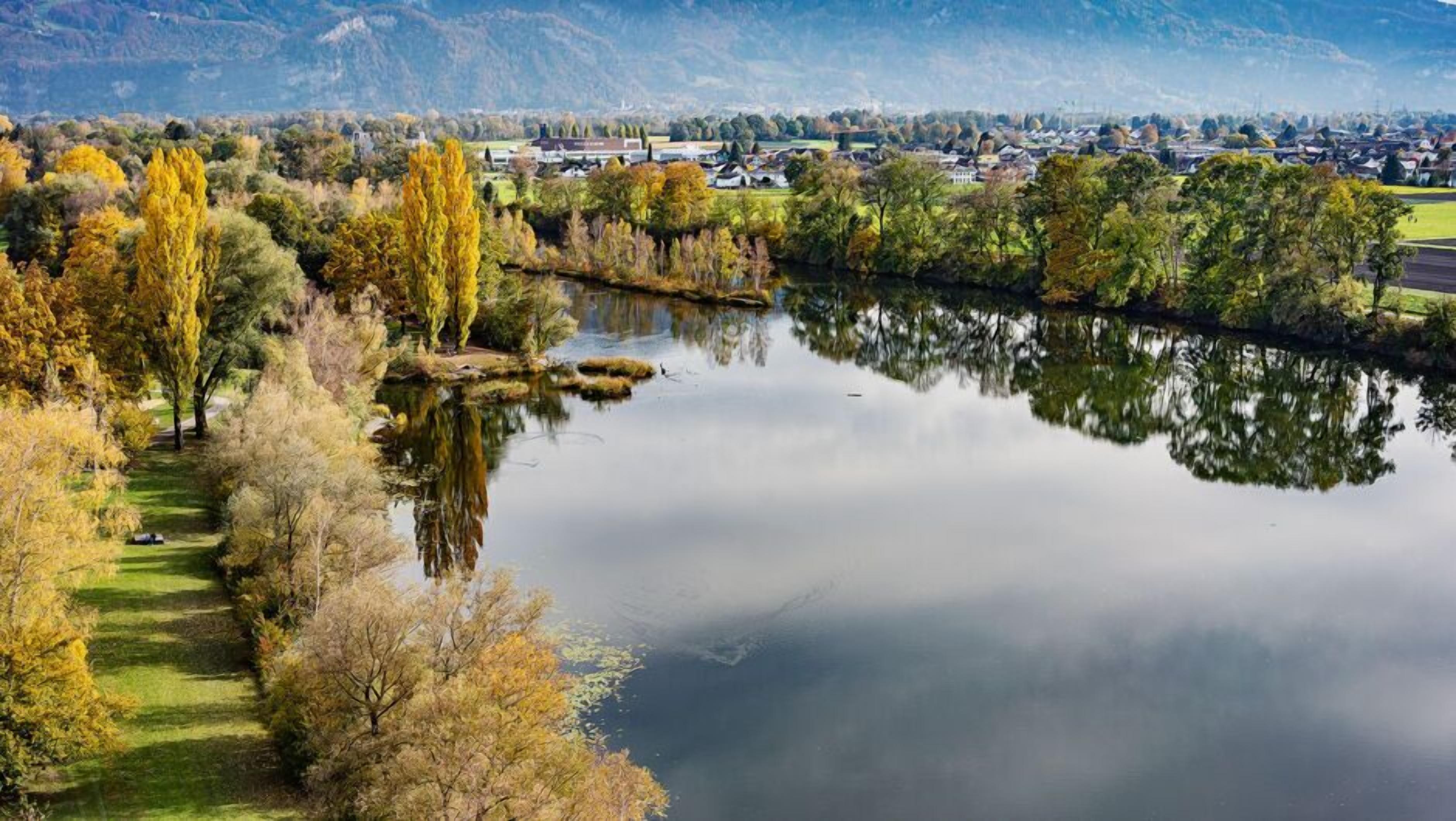 Um rio com árvores refletidas na água, uma trilha ao longo da margem e uma cidade ao fundo.
