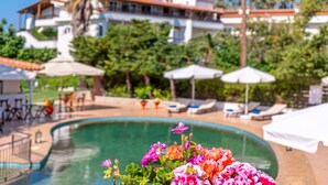 Uma área de piscina com espreguiçadeiras e guarda-sóis, cercada por vegetação e um edifício com telhado ladrilhado.