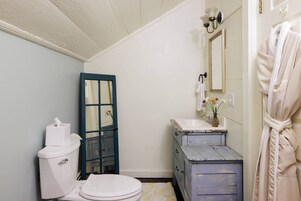 Casa de banho com sanita branca, porta azul, pia com um vaso de flores e um roupão pendurado à direita.