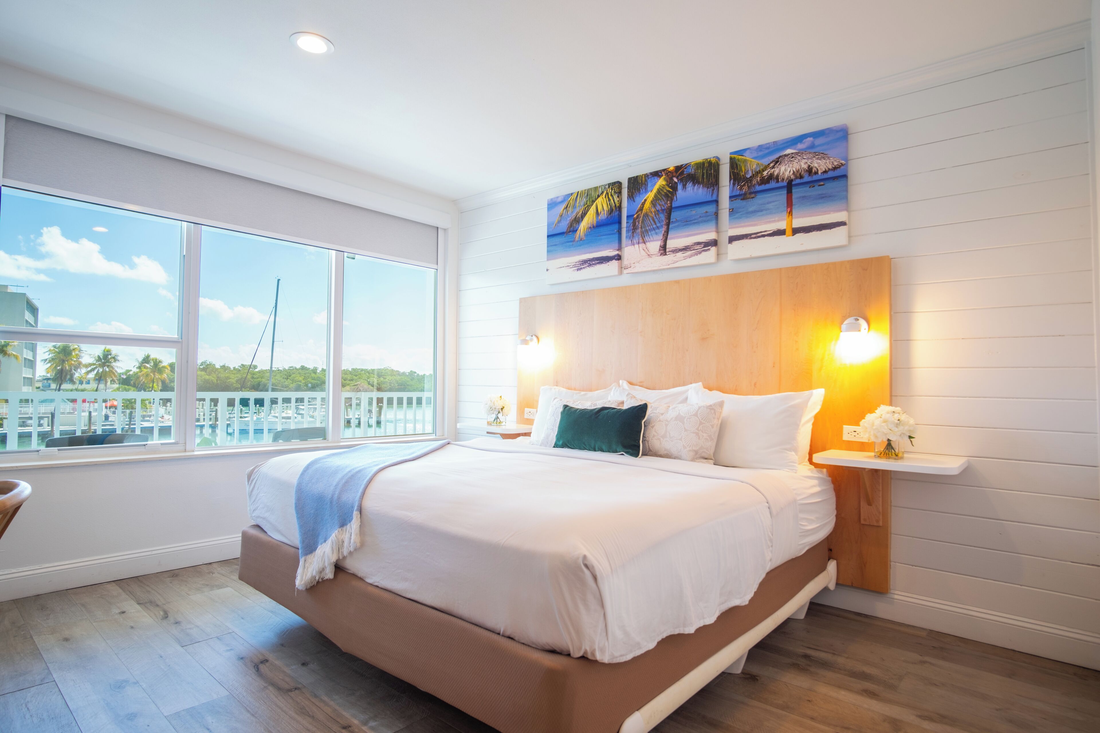 Quarto com cama grande, cabeceira de madeira e vista para a praia através de uma janela ampla.