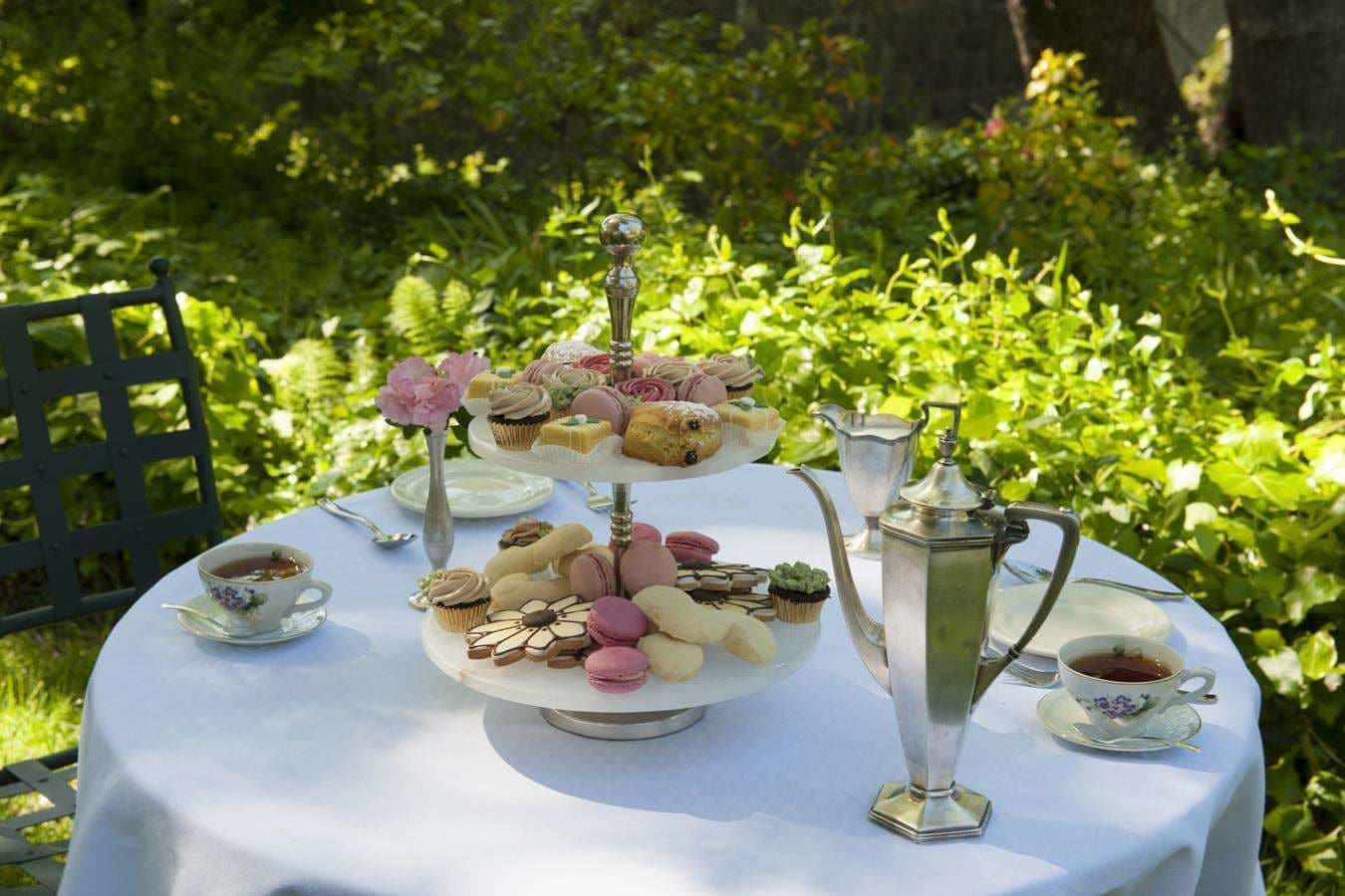 Suporte para sobremesas em três níveis, com diversos doces e biscoitos sobre uma toalha de mesa branca, acompanhado por um bule de prata e xícaras.