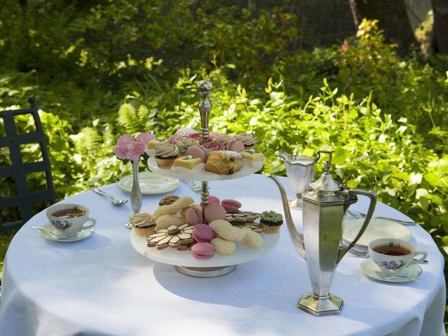 Suporte para sobremesas em três níveis, com diversos doces e biscoitos sobre uma toalha de mesa branca, acompanhado por um bule de prata e xícaras.