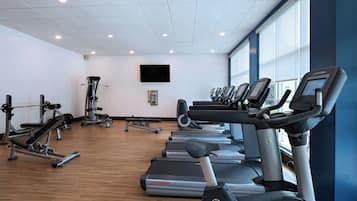 Academia com equipamentos de cardio, pesos e uma TV na parede.