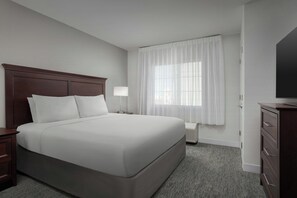 Quarto de hotel com uma cama grande, mesa de cabeceira, abajur, penteadeira e televisão.
