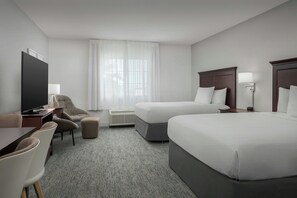 Quarto de hotel com duas camas, uma escrivaninha com televisão, uma cadeira, um sofá e uma janela grande com cortinas.