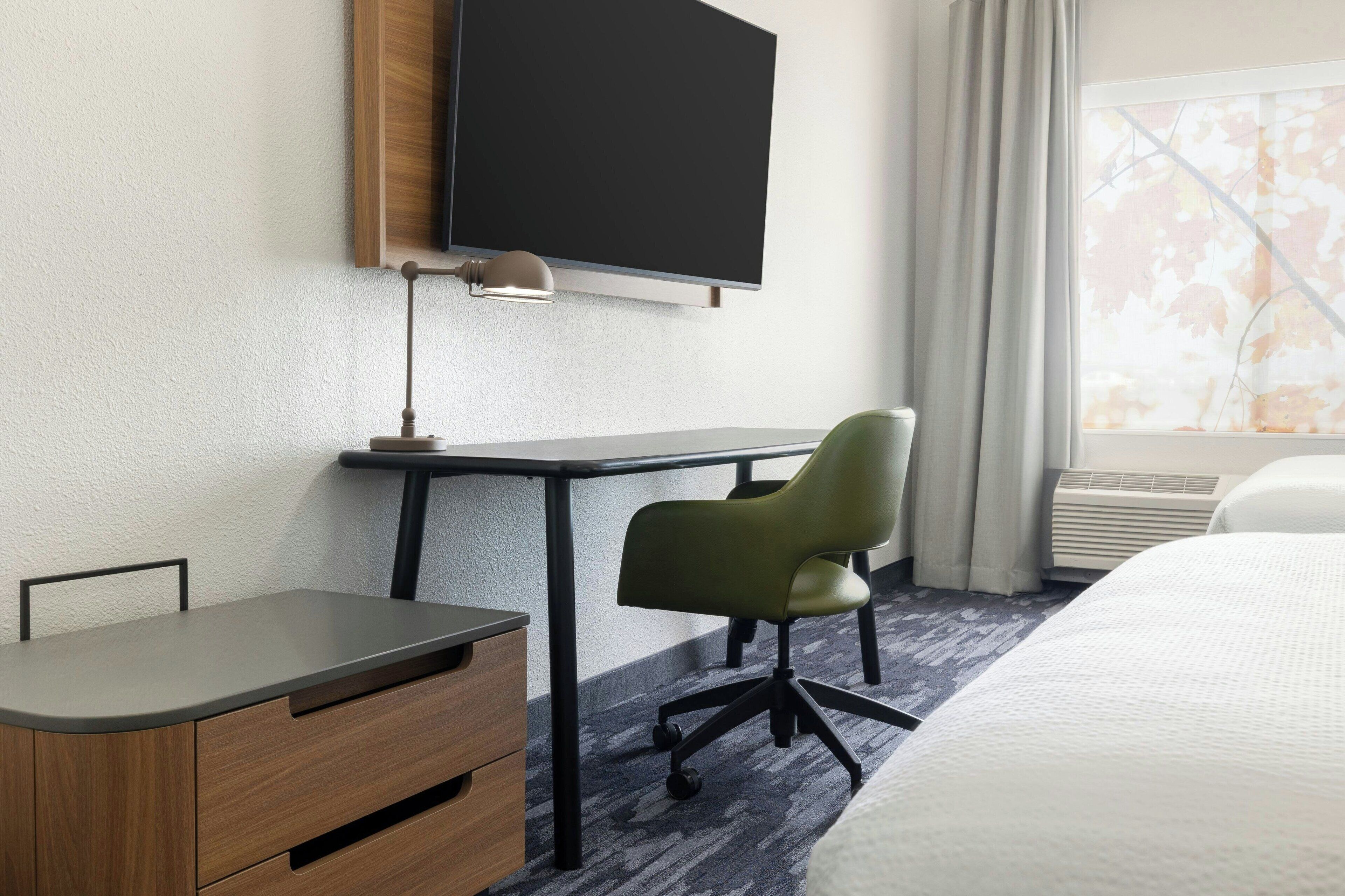 Quarto de hotel com cama, escrivaninha, cadeira, televisão e janela com cortinas.