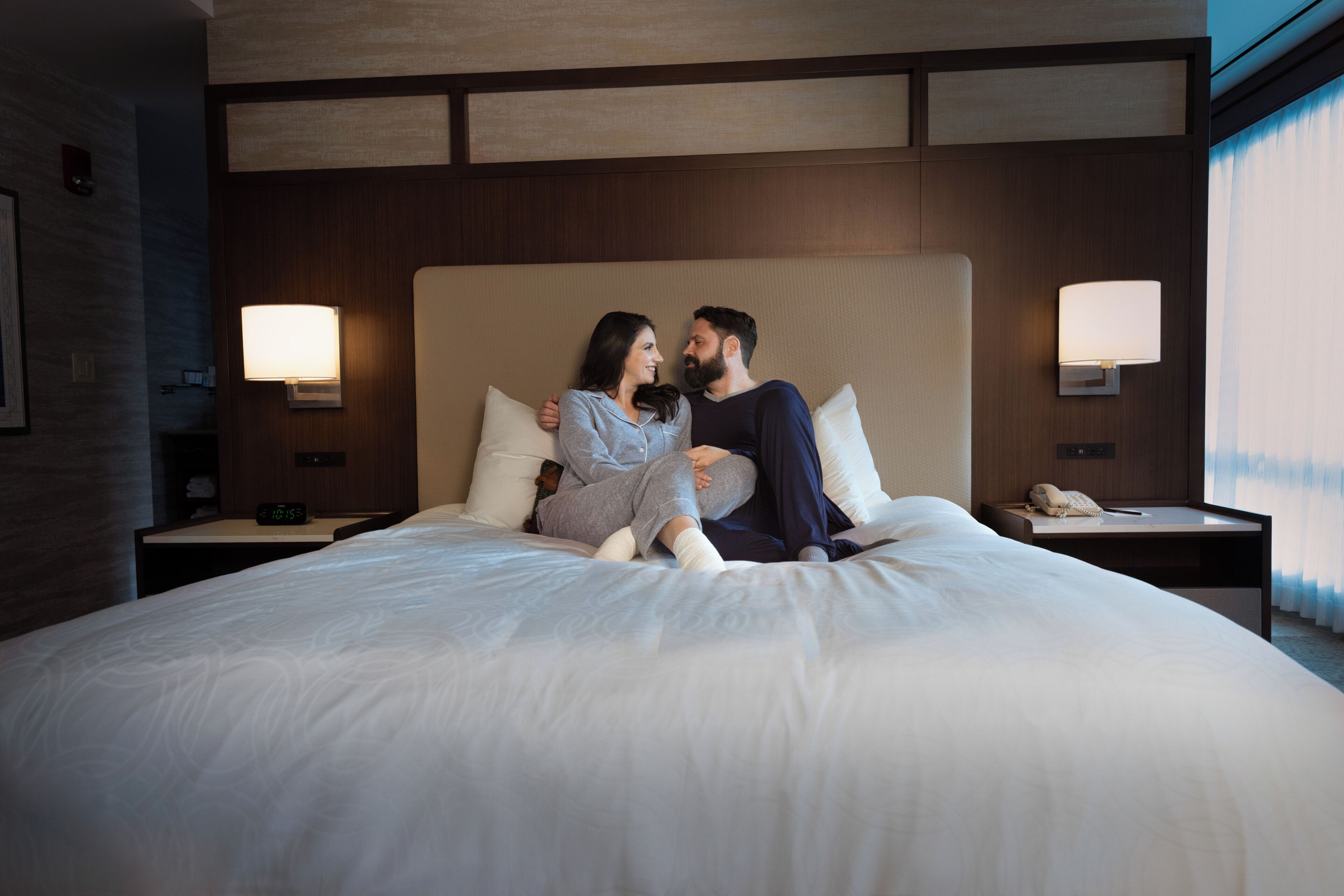 Quarto de hotel com uma cama grande, duas mesas de cabeceira, uma luminária na parede e um relógio sobre uma das mesas.