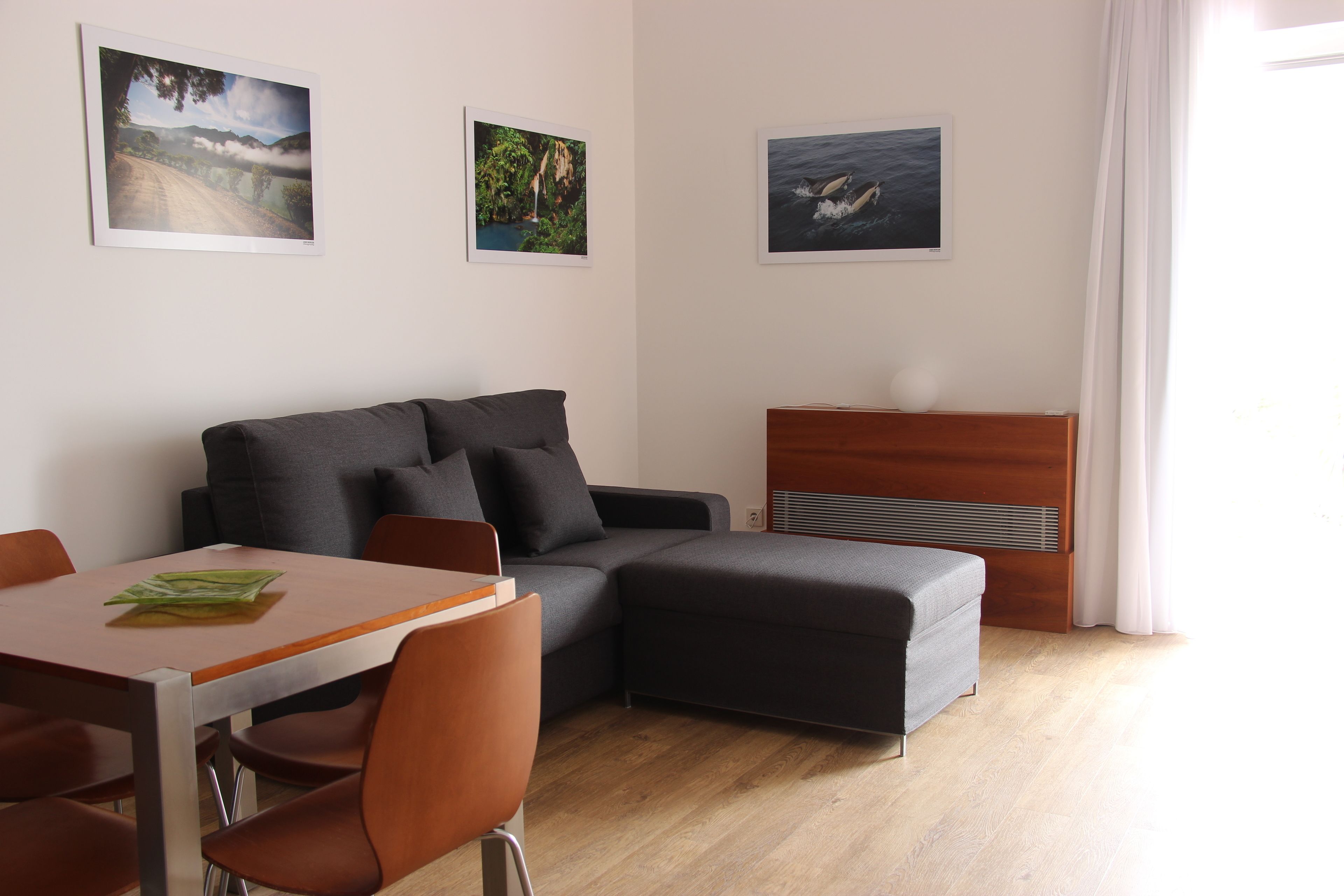 Uma sala de estar moderna com um sofá modular cinza, uma mesa de jantar de madeira com cadeiras laranja e uma cabeceira de madeira na parede.