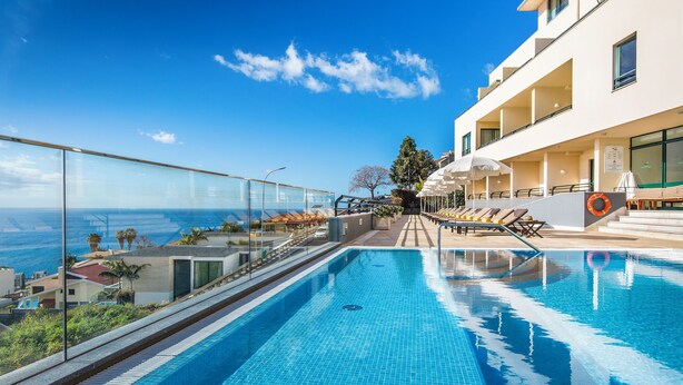 Céu azul e límpido, piscina com barreira de vidro e um edifício com varanda com vista para o mar.