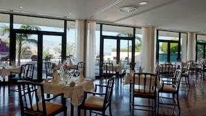 Um restaurante com várias mesas e cadeiras, grandes janelas com vista para palmeiras e uma paisagem distante.