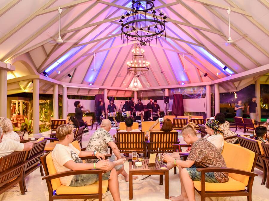 Uma grande estrutura em forma de tenda, com um candelabro, móveis de madeira e pessoas sentadas em mesas.