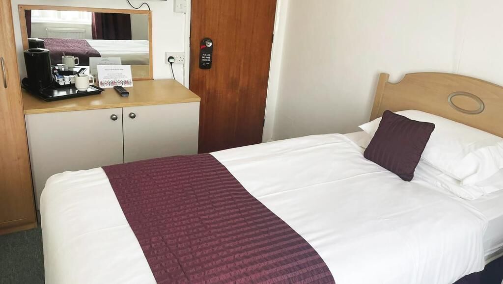 Quarto de hotel com cama, secretária de madeira com cafeteira, espelho e porta com placa.