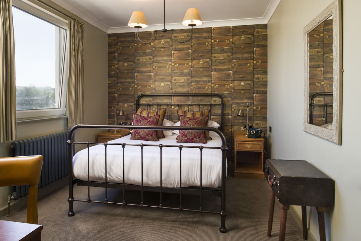 Quarto com cama de estrutura metálica, almofadas estampadas, mesa de cabeceira de madeira, mala e espelho na parede.
