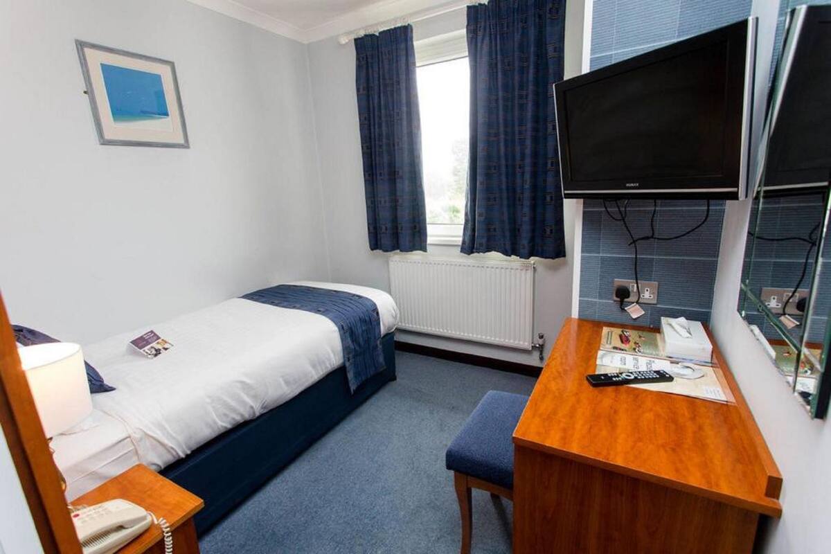 Quarto de hotel com uma cama de solteiro, uma escrivaninha com cadeira, televisão e janela com cortinas.