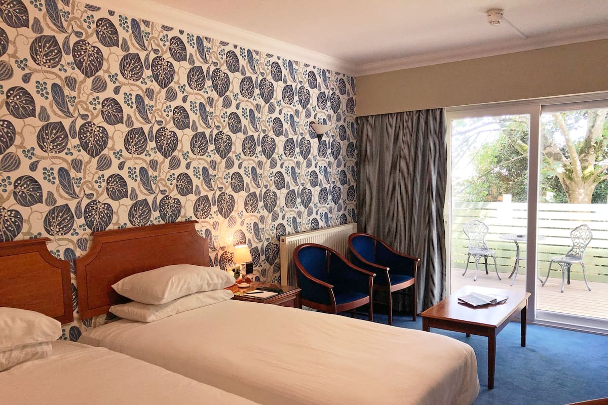 Quarto de hotel com duas camas, uma mesa pequena, duas cadeiras, um abajur e vista para o exterior através de uma porta deslizante.