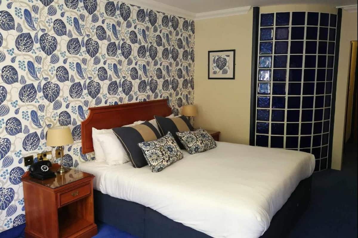 Uma cama bem arrumada com cabeceira de madeira, duas mesas de cabeceira com abajures e um papel de parede estampado.