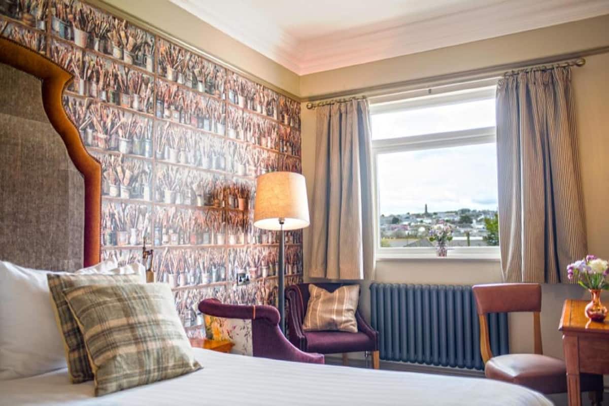 Quarto de hotel com uma cama grande, uma estante, uma janela com vista e uma cadeira.
