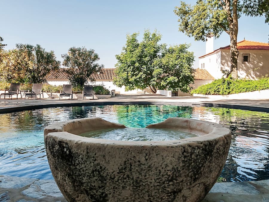 Uma fonte de pedra em uma piscina, com espreguiçadeiras e um edifício ao fundo.