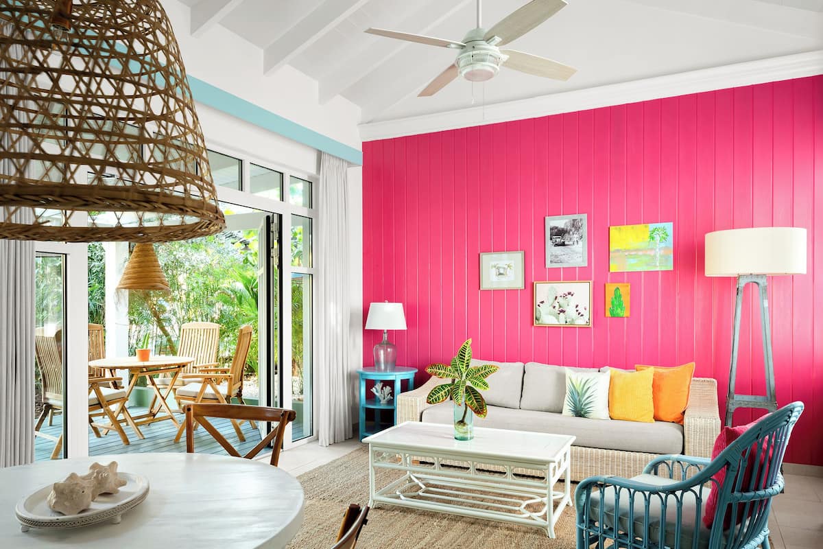 Uma sala de estar luminosa com parede rosa, um ventilador de teto e um espaço de refeições com móveis de vime.