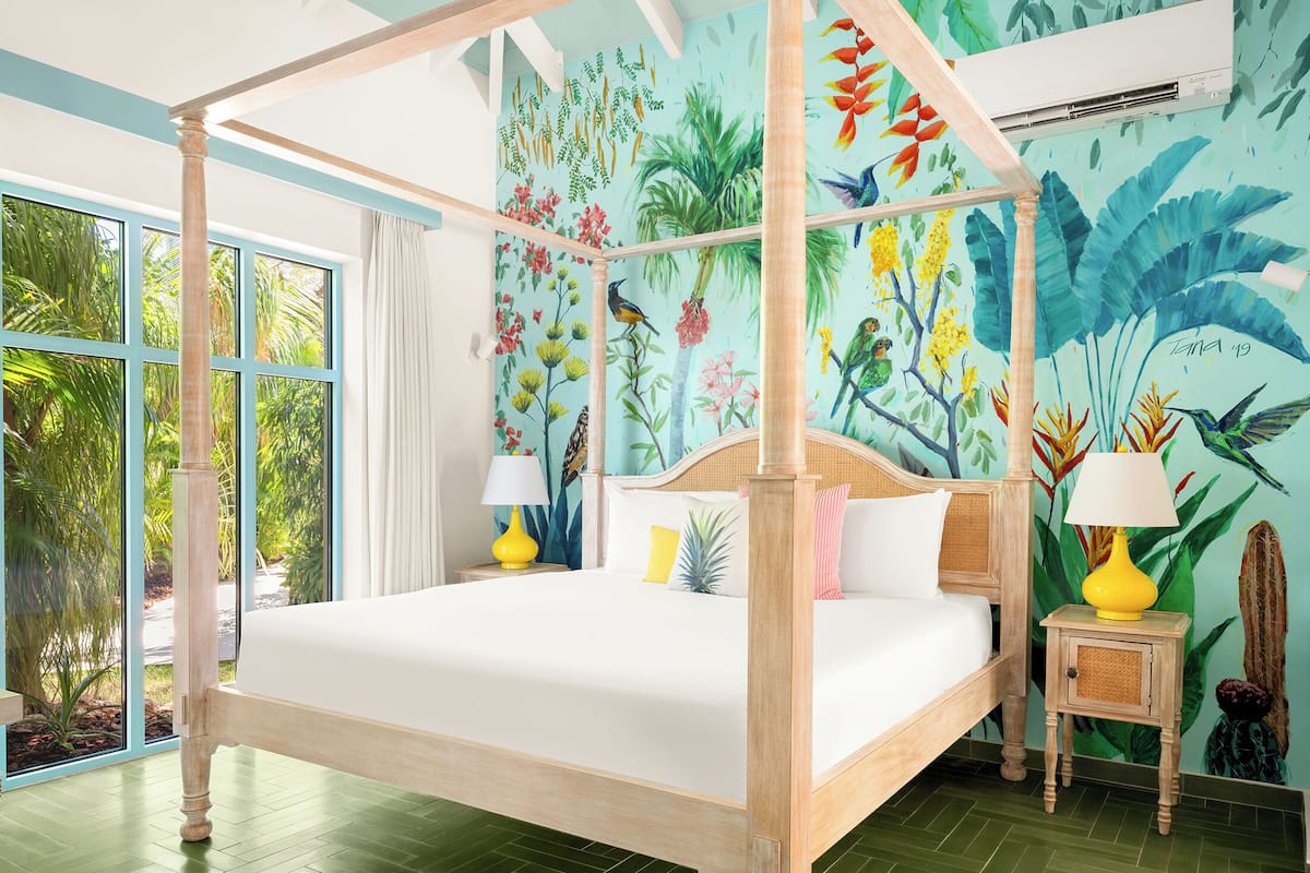 Uma cama de dossel com estrutura de madeira, colchão branco e cabeceira com encosto trançado. Há duas mesas de cabeceira com luminárias amarelas, e uma janela grande com cortinas no lado esquerdo. O quarto apresenta um mural tropical vibrante na parede.
