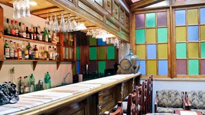 Bar com prateleiras de madeira repletas de garrafas de bebidas, copos pendurados e uma grande janela com painéis coloridos.