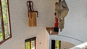 Quarto com parede branca texturizada, uma cadeira de madeira, uma escultura pendurada e uma janela com vista para o verde.