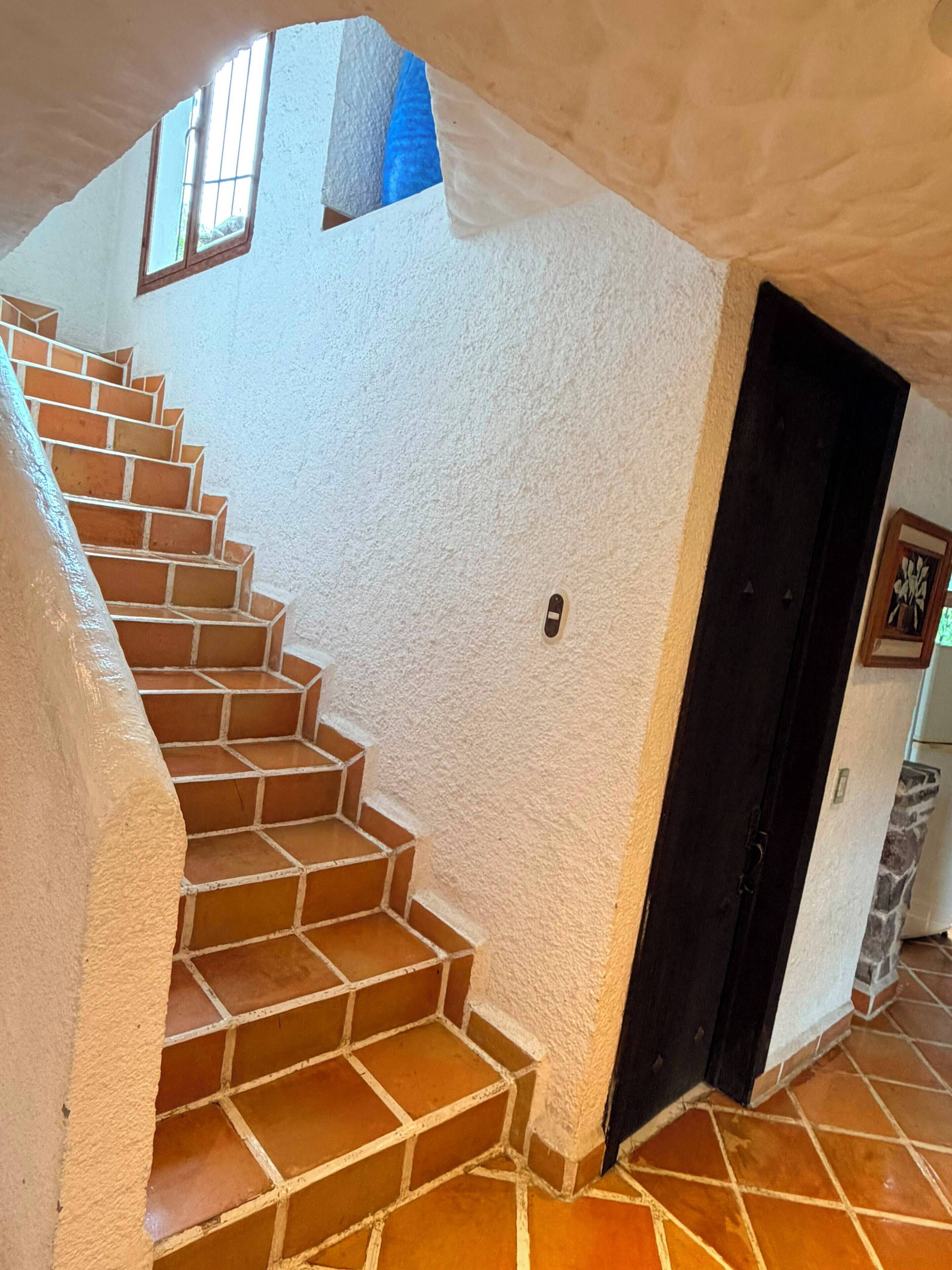 Uma escadaria com ladrilhos de barro que leva a uma porta, paredes brancas com textura e uma janela.