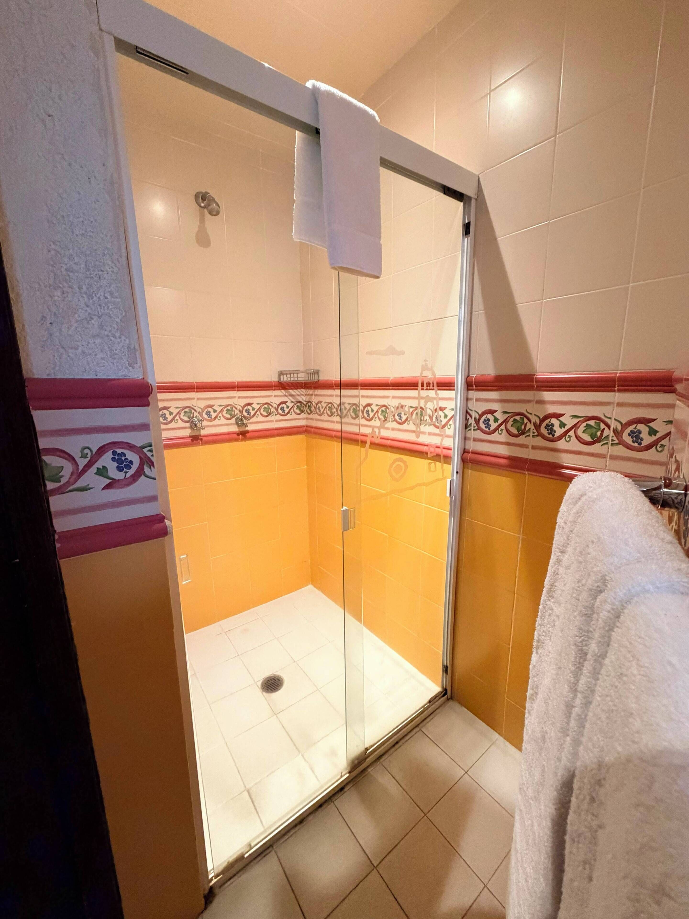Um chuveiro com paredes de vidro, uma toalha branca pendurada na porta, paredes revestidas com uma borda decorativa e um ralo visível no chão.