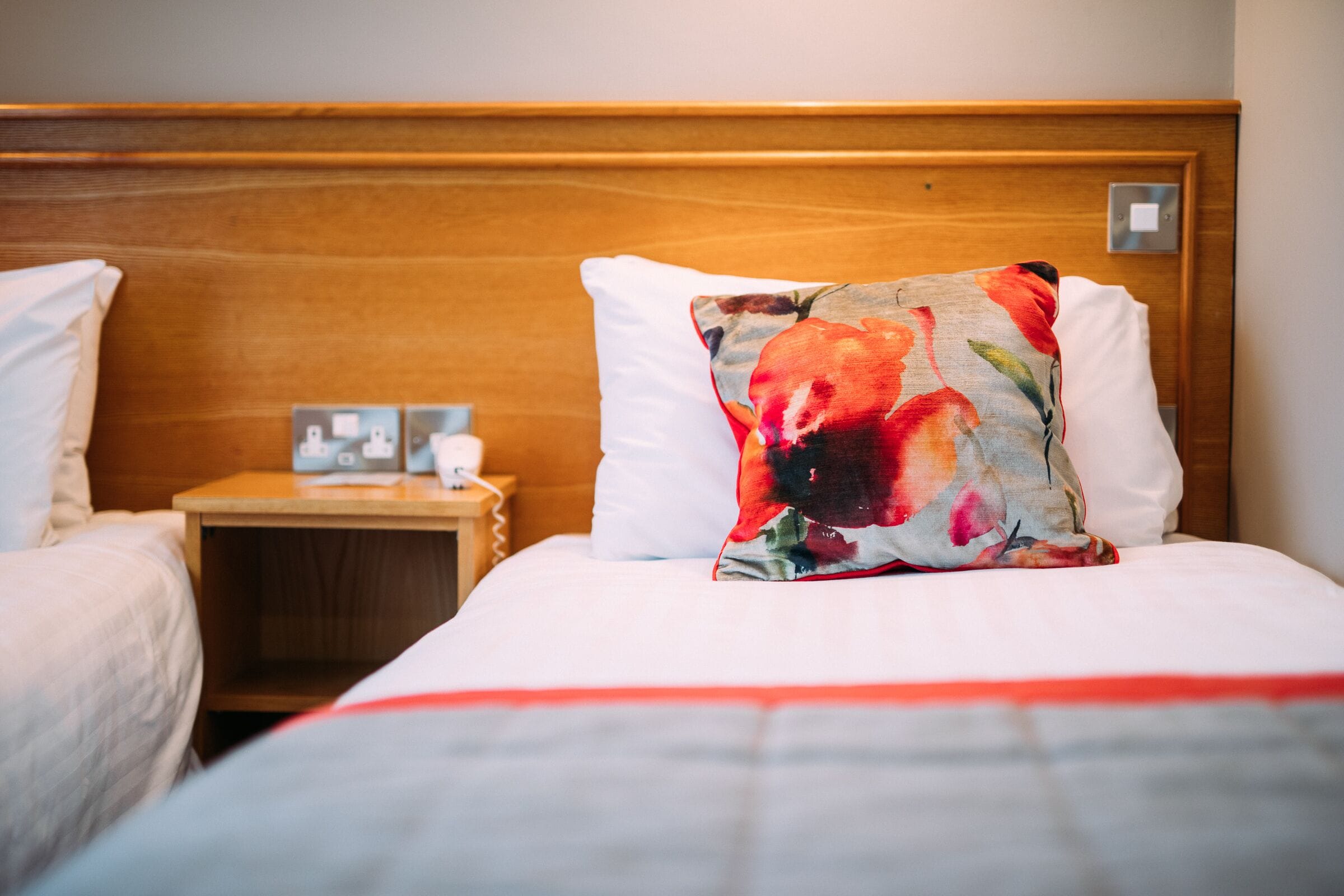 Quarto de hotel com cabeceira de madeira, cama com roupa de cama branca e um toque de vermelho, mesa de cabeceira com abajur e almofada floral.