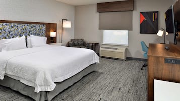 Quarto de hotel com uma cama grande, uma escrivaninha com computador, uma televisão e uma área de estar com uma cadeira e uma mesa pequena.