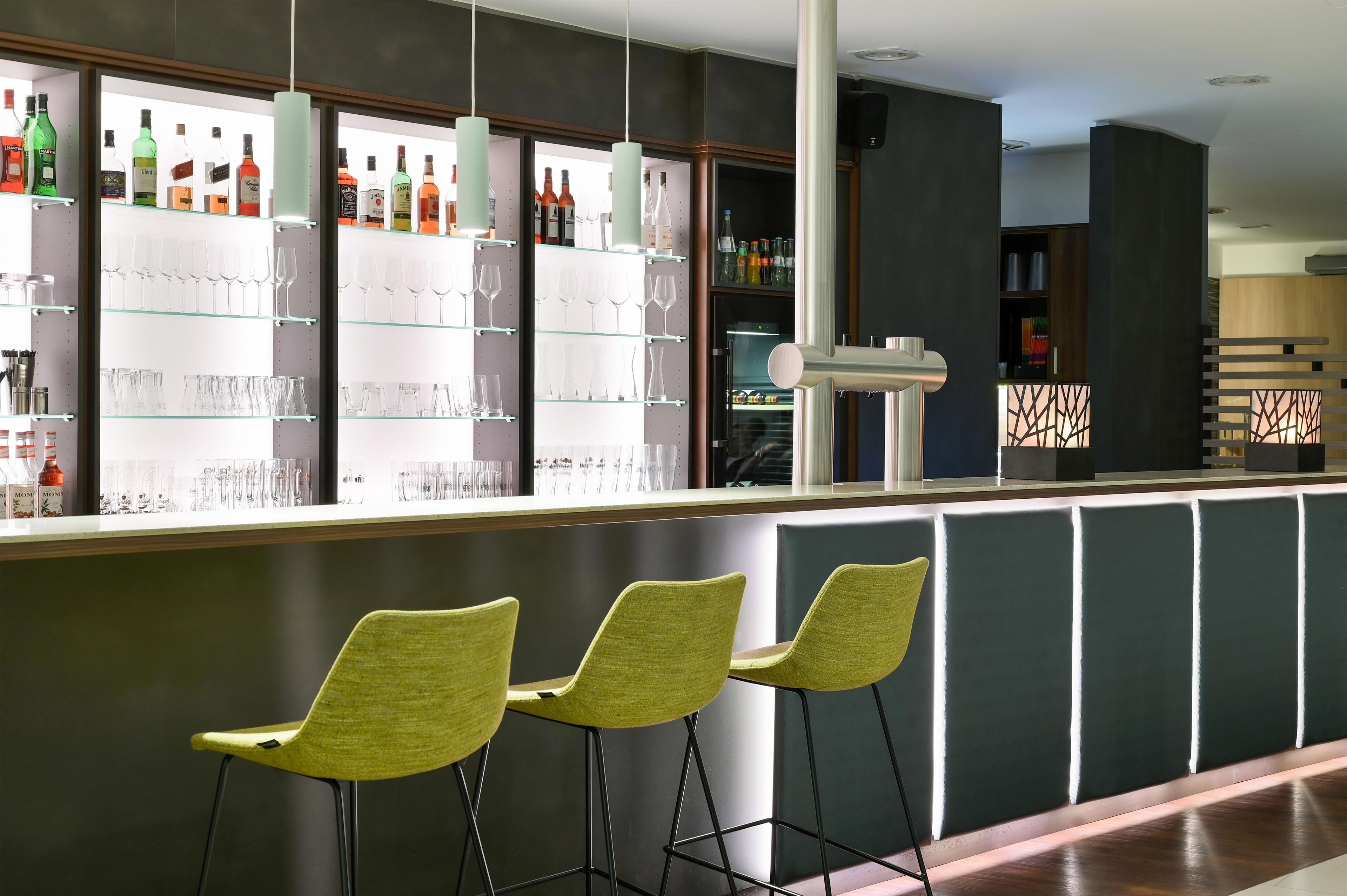 Uma área de bar com balcão, prateleiras com garrafas, copos e uma geladeira. Há três cadeiras verdes em frente ao bar.