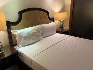 Cama bem arrumada com roupa de cama branca, cabeceira de madeira e duas luminárias de mesa.