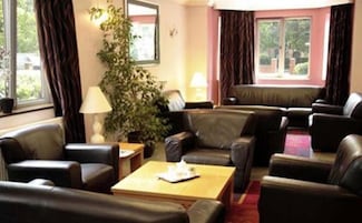 Quarto de hotel com sofás de couro, uma mesa de centro, luminária e uma janela grande com cortinas.