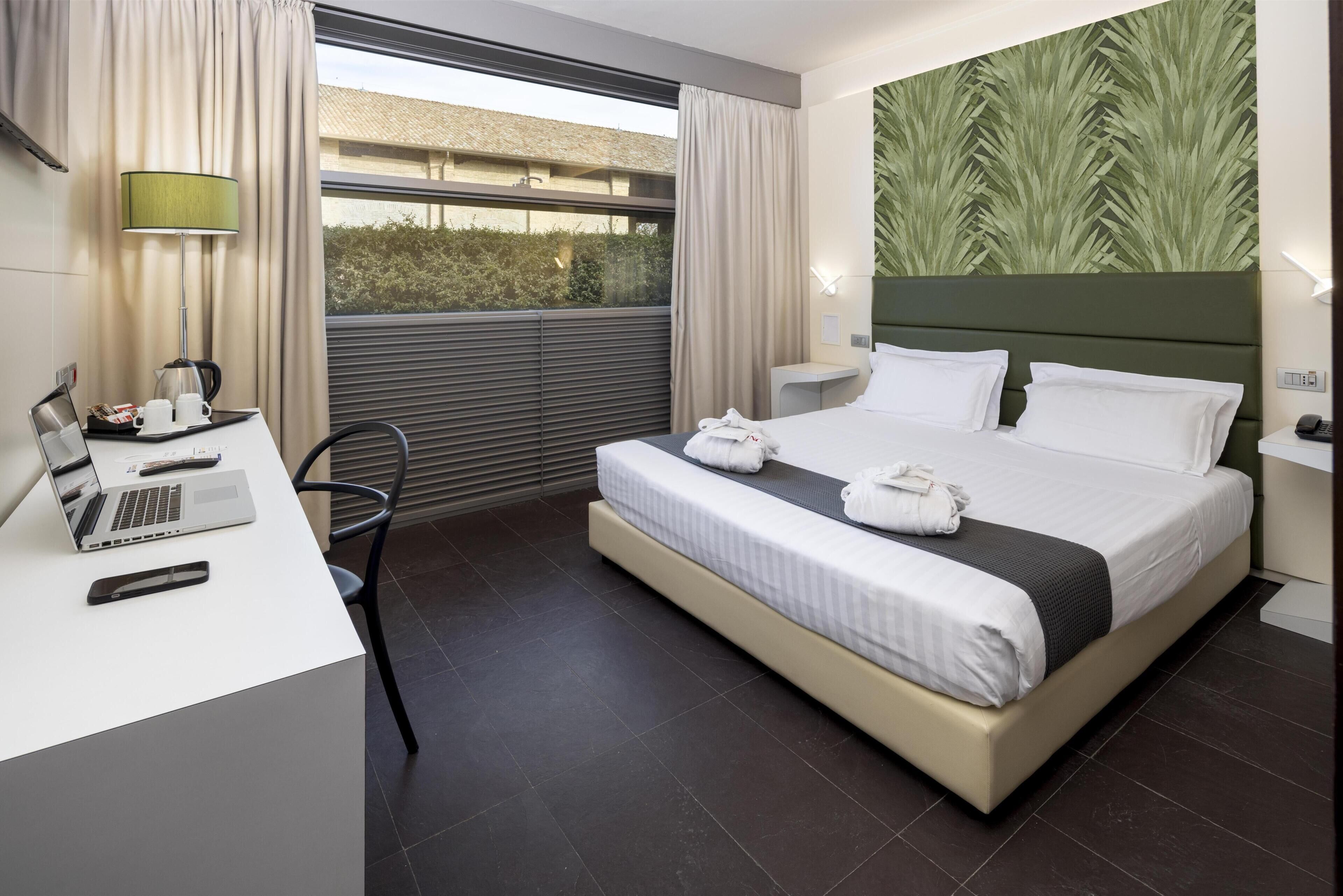 Quarto de hotel com cama, escrivaninha com um laptop, telefone e janela com cortinas.