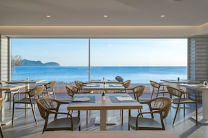 Um restaurante com mesas e cadeiras, grandes janelas com vista para o mar.