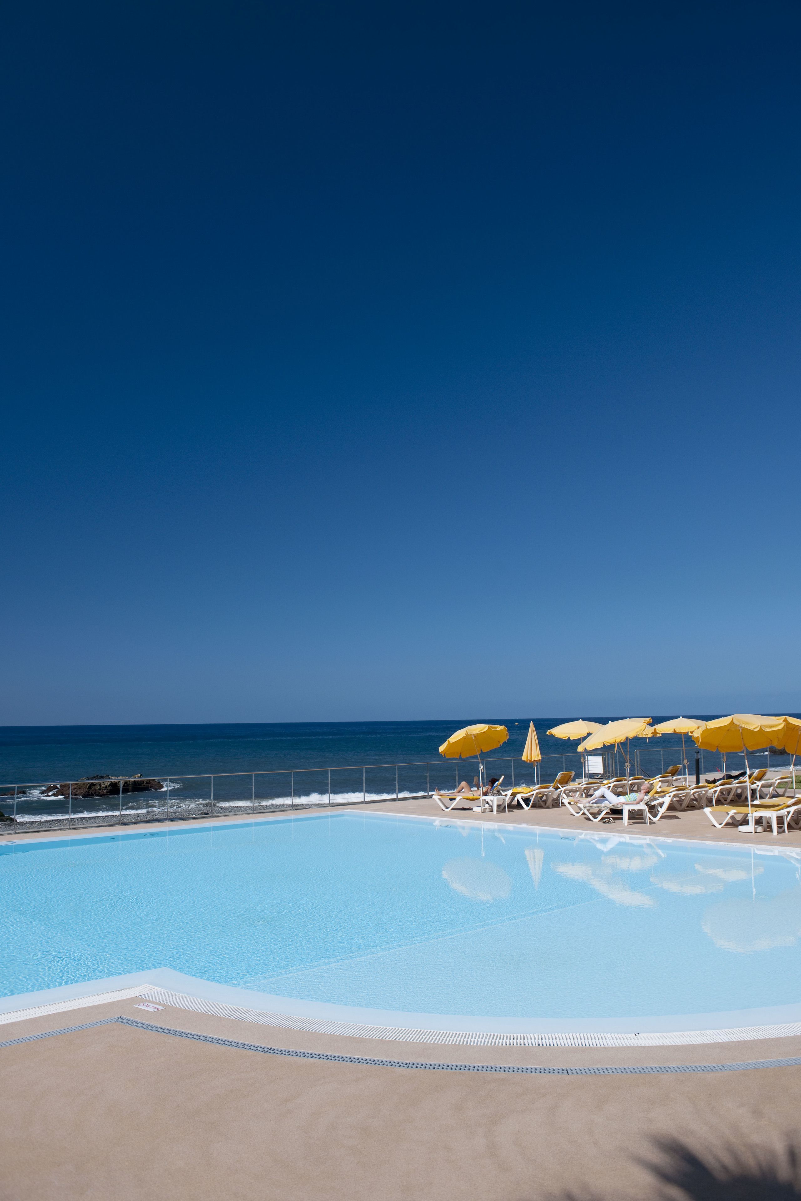 Céu azul límpido, piscina com espreguiçadeiras e guarda-sóis amarelos, e uma linha costeira rochosa ao fundo.