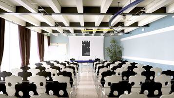 Uma sala de conferências com fileiras de cadeiras voltadas para uma tela de apresentação, uma planta à direita e uma escada ao fundo.