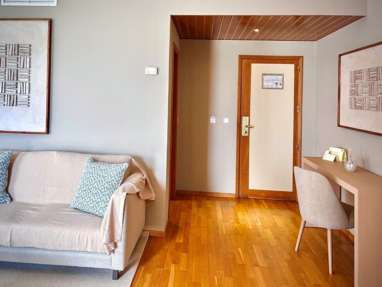 Um quarto com piso de madeira, um sofá com almofadas estampadas, uma escrivaninha com cadeira e uma porta com uma placa.