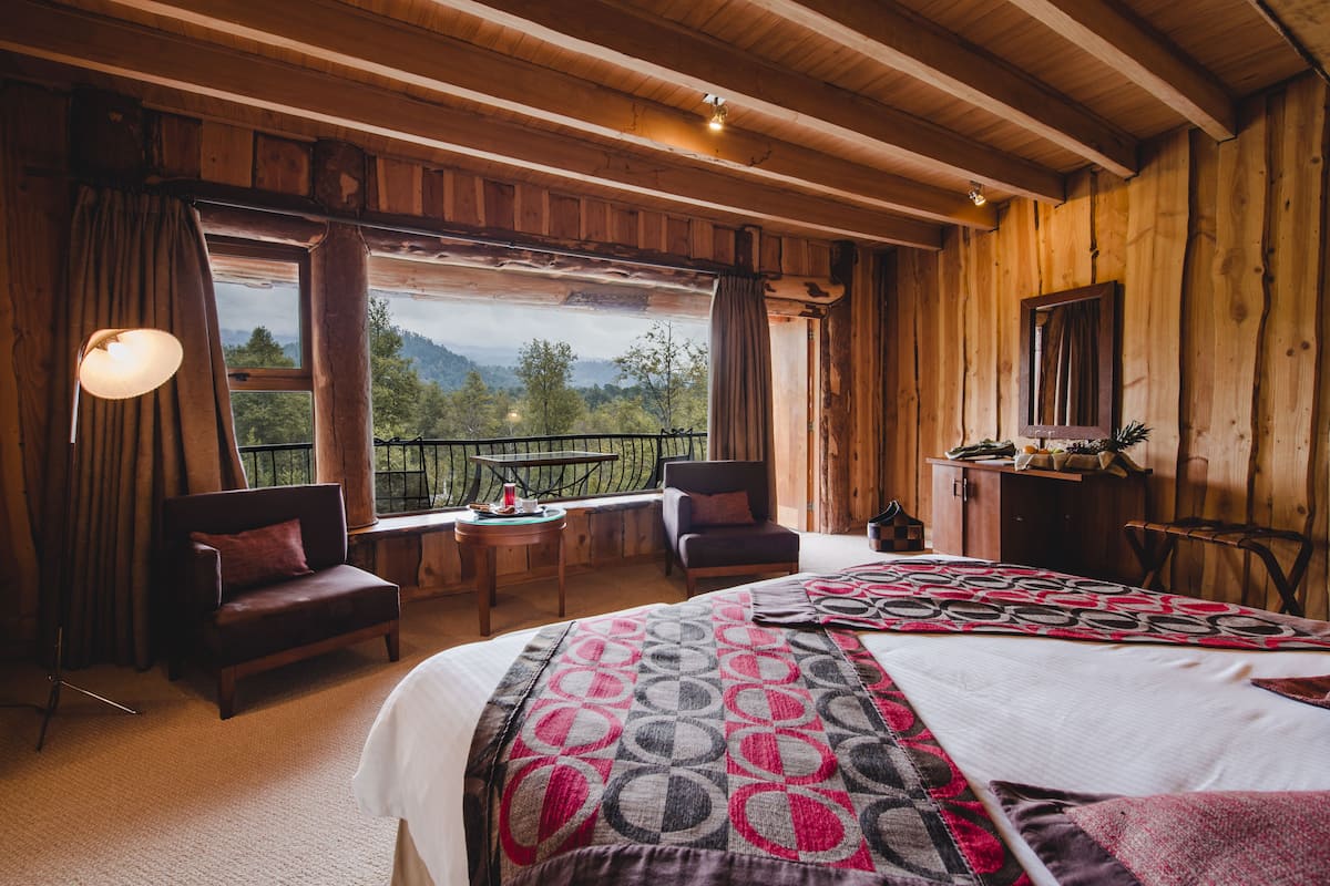 Quarto de cabana de madeira com uma cama grande, duas cadeiras, uma mesa e uma varanda com vista para as montanhas.