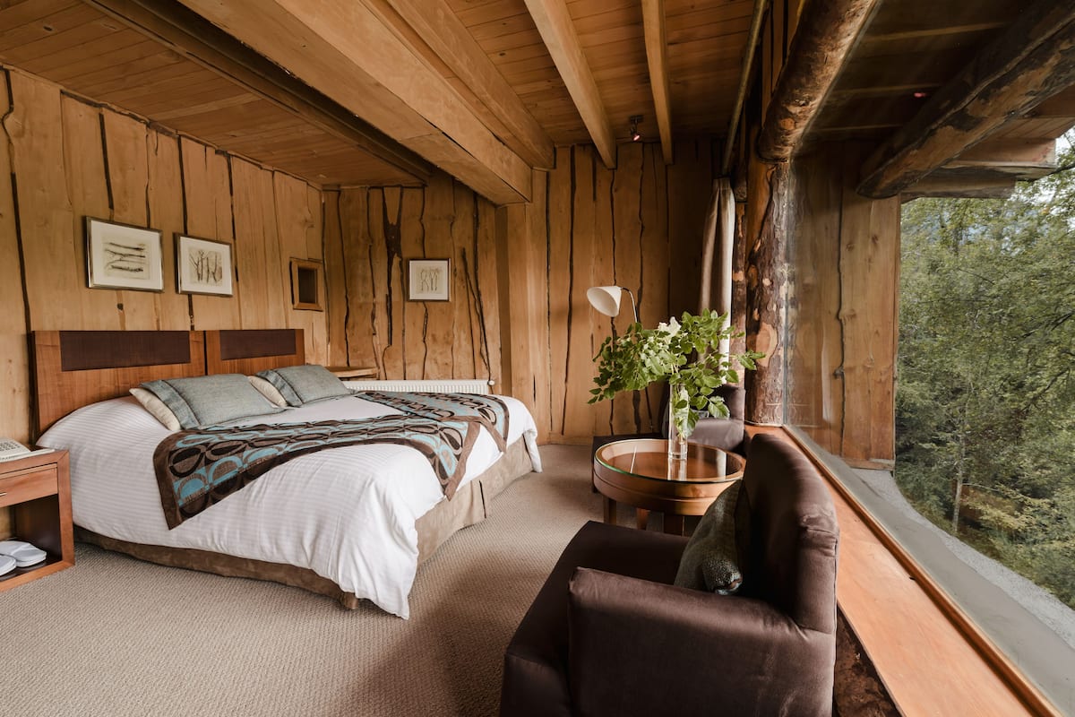 Quarto de cabana de madeira com uma cama grande, uma mesa de cabeceira, uma cadeira e vista para a floresta.