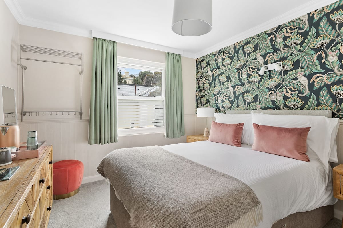 Quarto com uma cama grande, cômoda de madeira, um banquinho vermelho, janela com cortinas verdes e papel de parede floral.